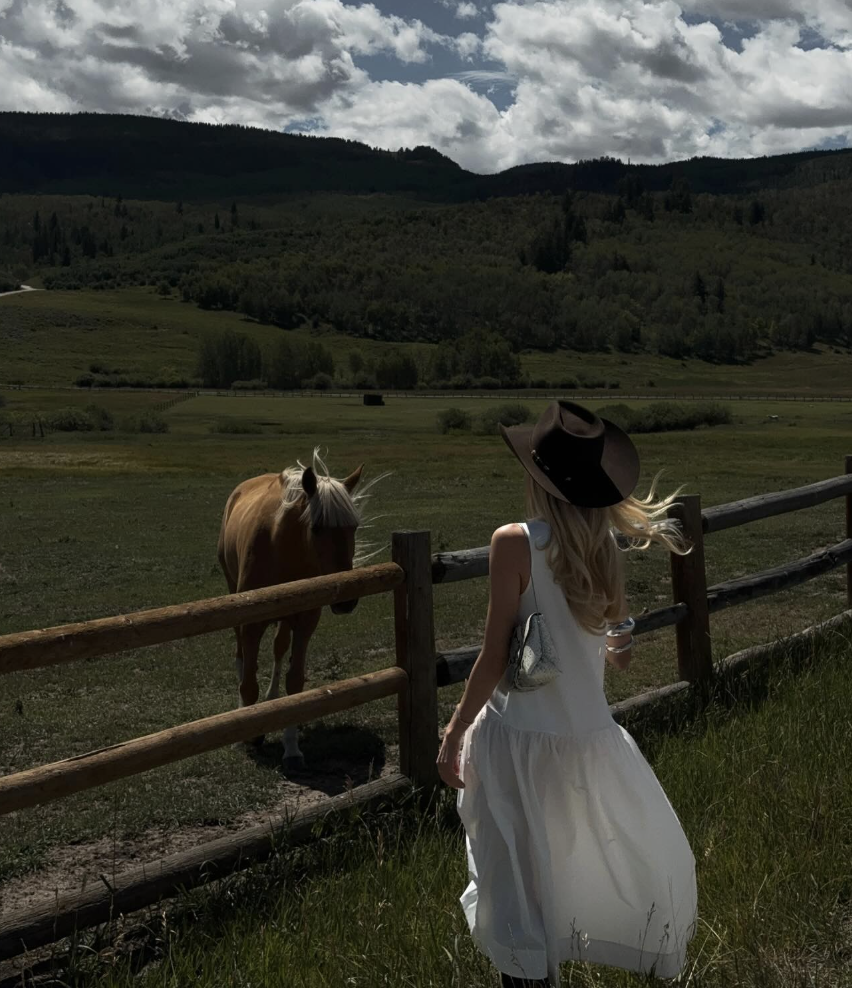 Woman wearing a flowing white sleeveless midi dress with a black wide-brim hat and silver handbag in a countryside setting with a horse and wooden fence.